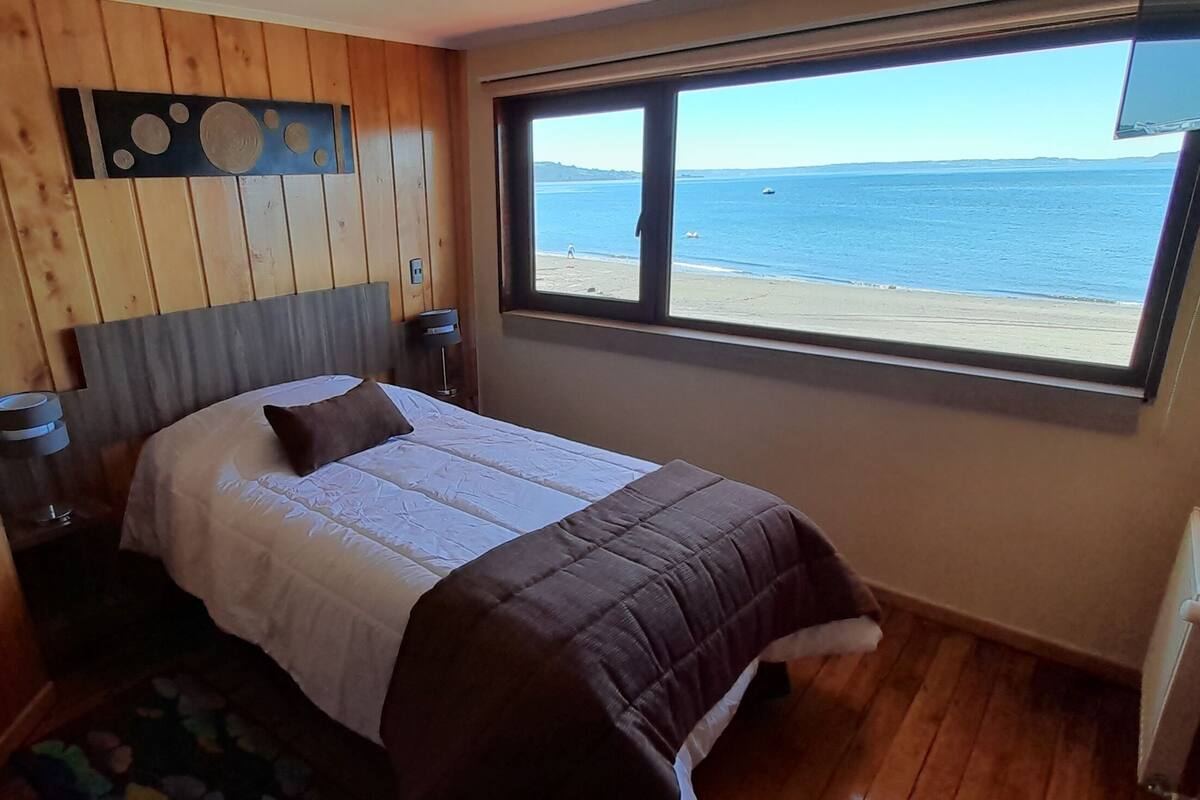 Quarto com cama, paredes de madeira, janela vista para a praia e televisão na parede.