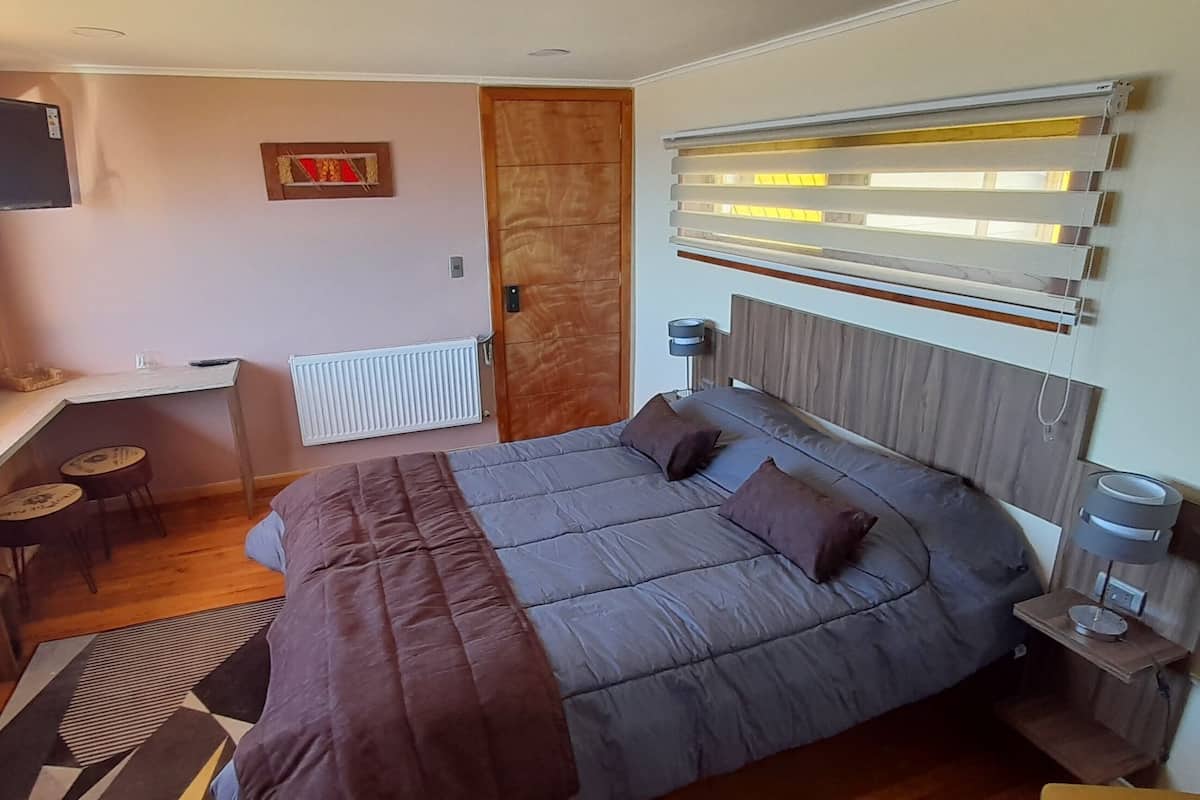 Quarto com uma cama grande, mesa de cabeceira com abajur, janela com persianas, radiador e TV fixada na parede.