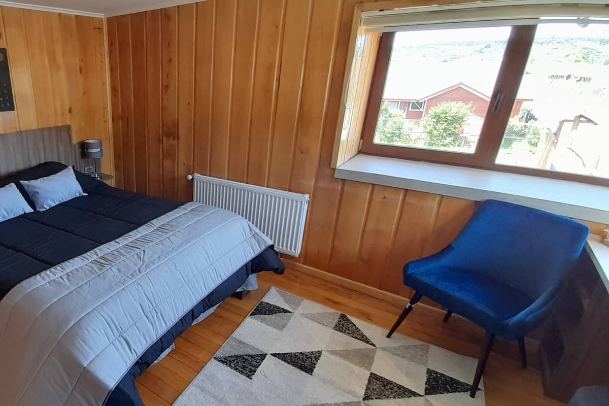 Quarto com parede de painel de madeira, cama com edredom cinza e branco, cadeira azul e janela com vista para casas.