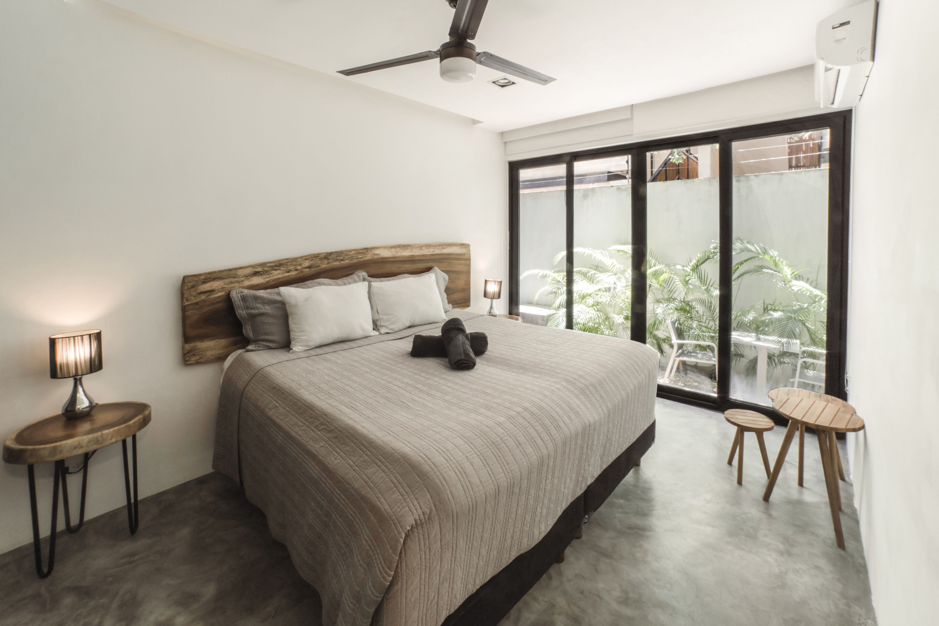 Quarto com cama grande, ventilador de teto, uma mesinha e vista para o exterior.