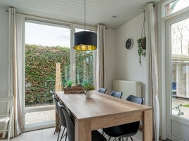 Uma sala de jantar com mesa de madeira, cadeiras pretas, luminária pendente e vista para um jardim através de grandes janelas.
