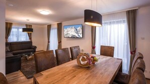 Quarto de hotel moderno com mesa de jantar de madeira, cadeiras de couro, sofá e uma janela grande com cortinas.