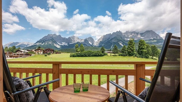 Um deck de madeira com uma mesa redonda e duas cadeiras, com vista para uma paisagem montanhosa e edifícios no vale.