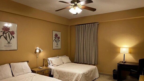 Quarto de hotel com duas camas, uma escrivaninha, uma cadeira, um ventilador de teto e duas luminárias.