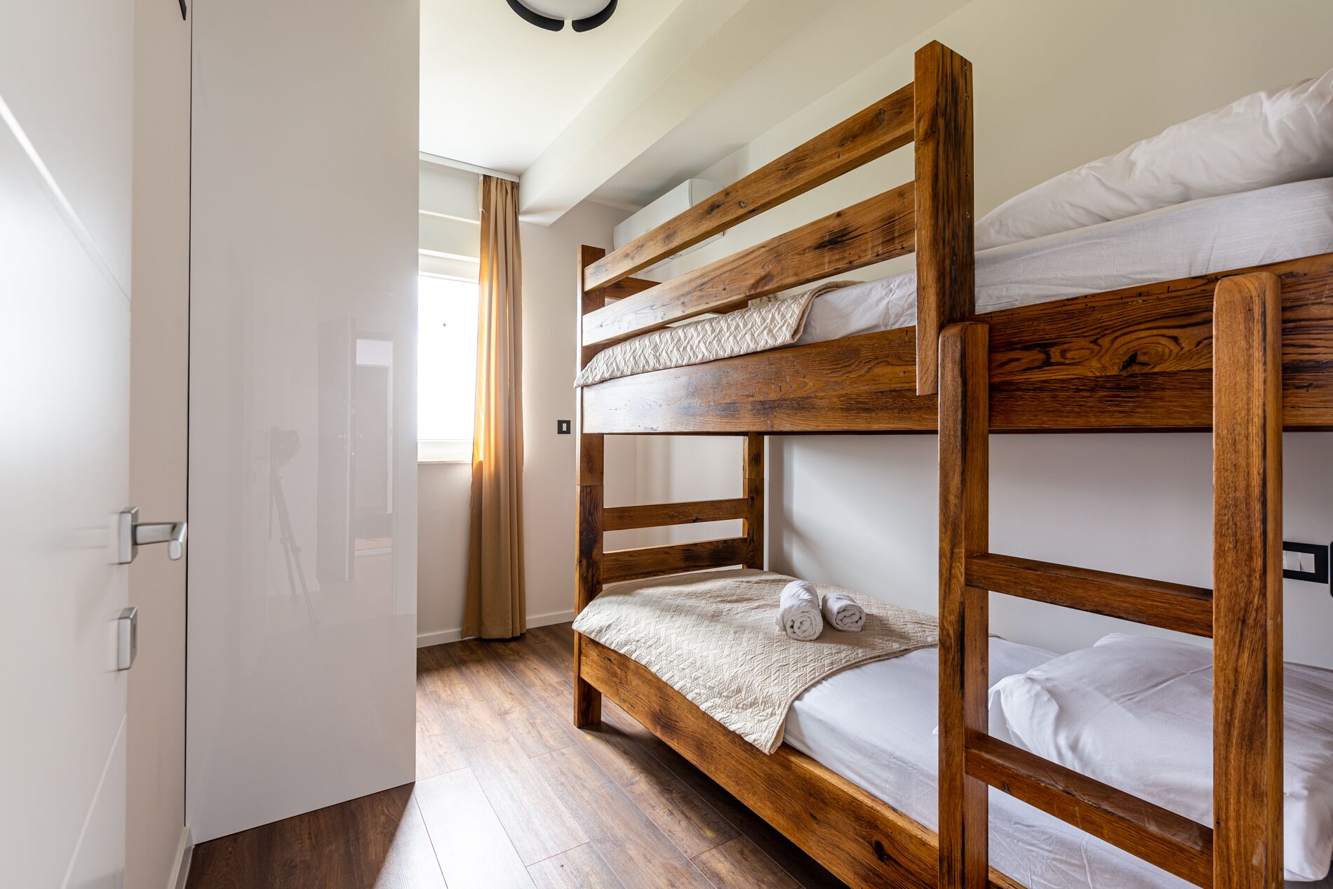 Quarto com beliches de madeira, roupa de cama branca e porta que dá para outro cômodo.