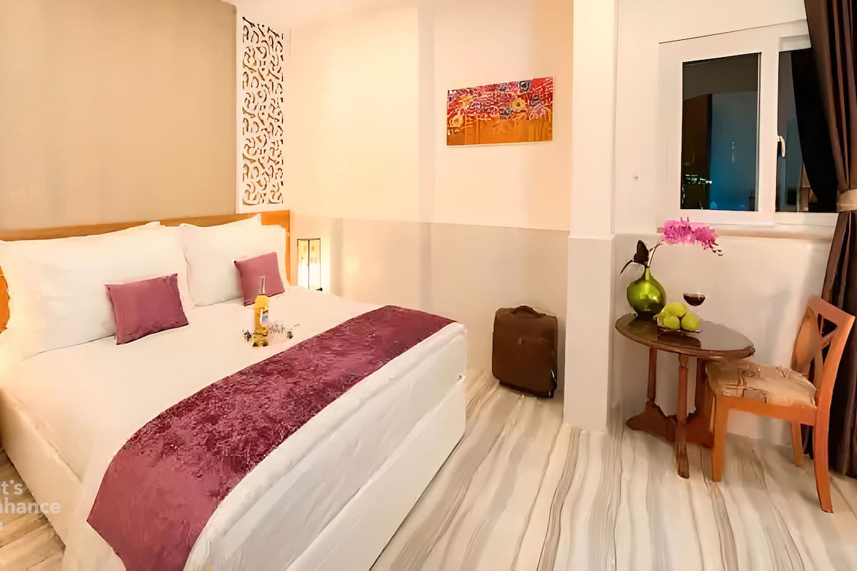 Quarto de hotel com cama, uma mesinha com um vaso e frutas, uma cadeira e uma janela com cortinas.