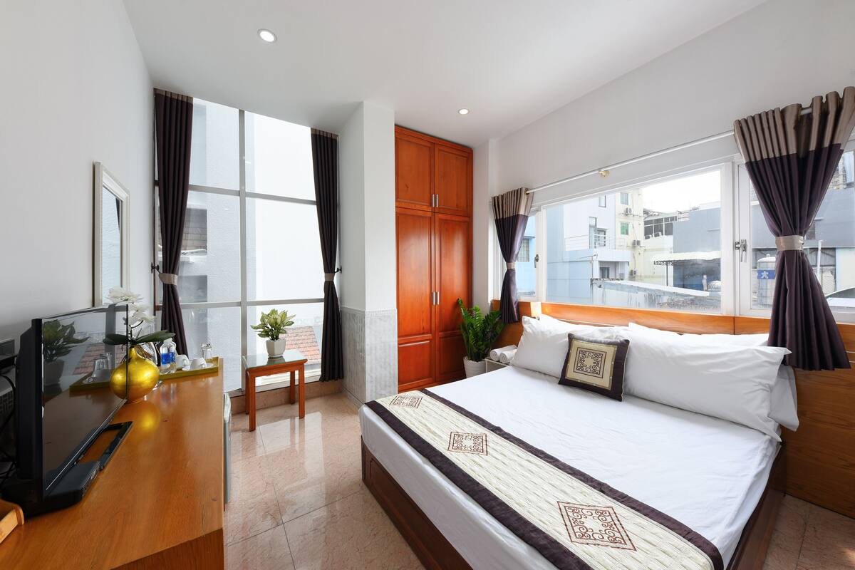 Quarto de hotel com uma cama grande, guarda-roupa de madeira, uma escrivaninha com computador, janela com cortinas e vista para edifícios.