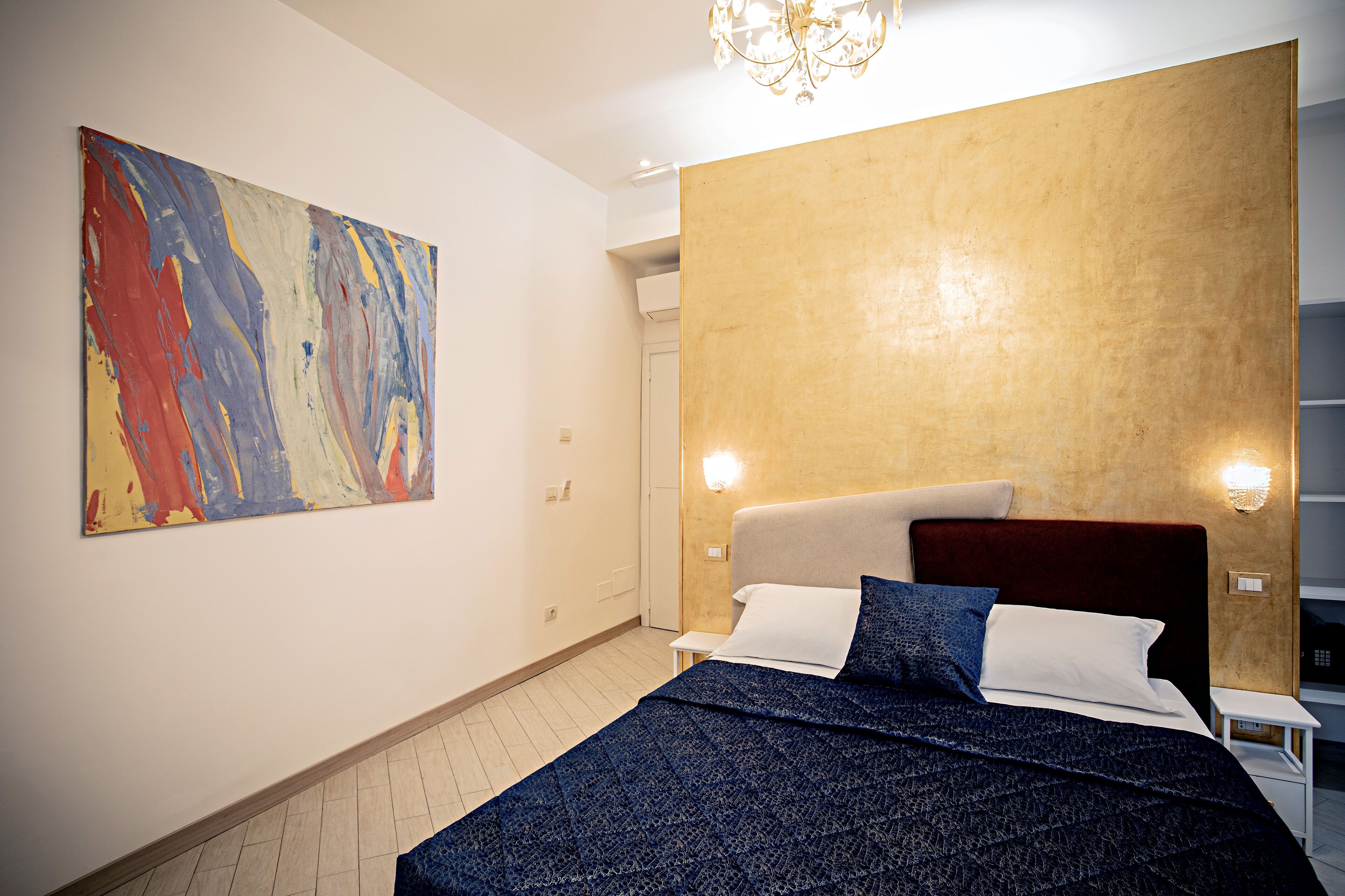 Quarto moderno com cama grande, pintura abstrata colorida, cabeceira de madeira e mesa de cabeceira pequena.