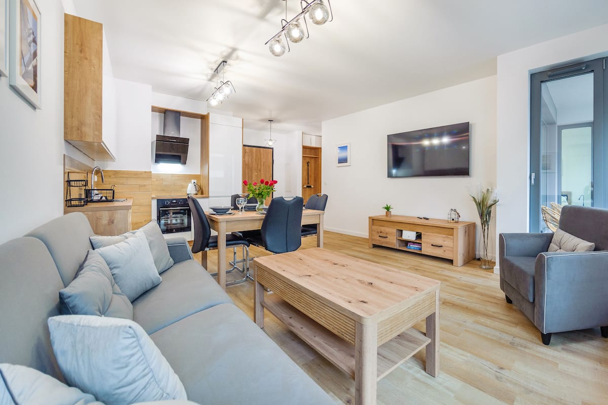 Uma sala de estar moderna com sofá, mesa de centro e televisão. Há uma área de jantar com mesa e cadeiras. O ambiente possui piso de madeira e paredes brancas.