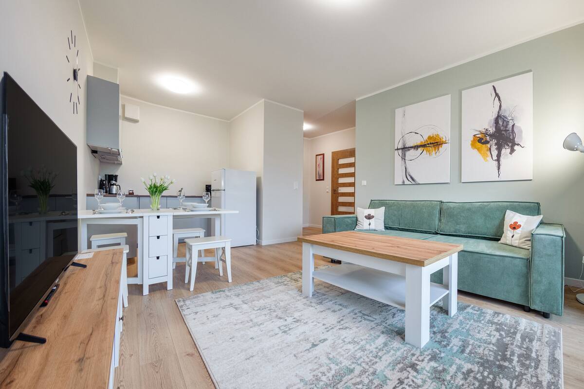 Uma sala de estar moderna com uma mesa de centro de madeira, um sofá verde e uma área de cozinha com uma geladeira branca.