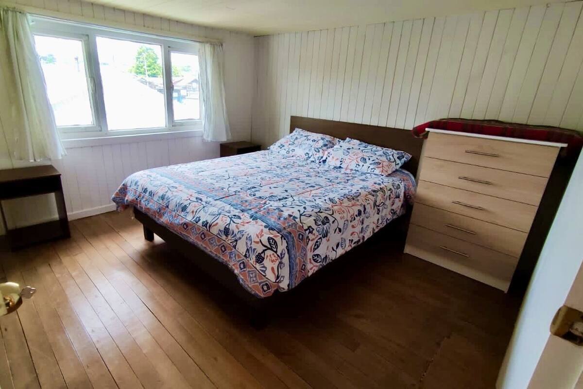 Um quarto com piso de madeira, uma cama com colcha florida, cômoda, janela com cortinas e porta.
