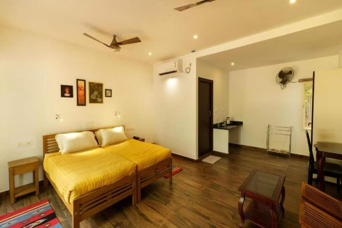 Quarto com cama, mesa de cabeceira e uma pequena escrivaninha com cadeira. Há um ventilador de teto, um aparelho de ar condicionado e um ventilador fixado na parede.