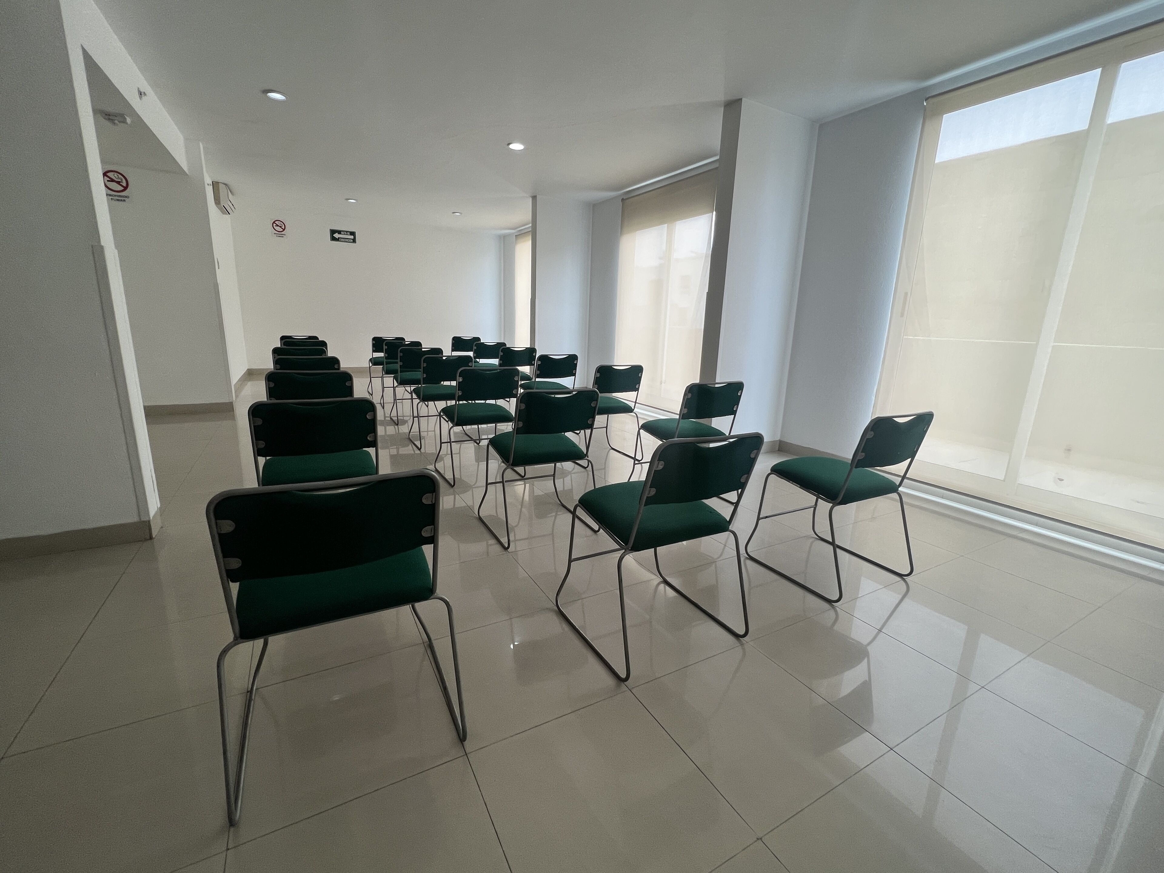 Uma sala de conferências com fileiras de cadeiras verdes, janelas amplas e piso de azulejos.