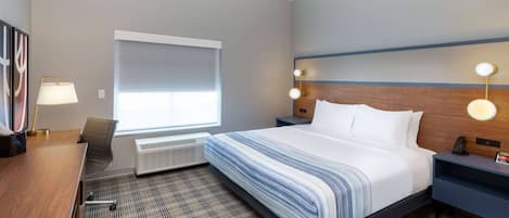 Quarto de hotel com uma cama grande, uma escrivaninha e uma cadeira. Há uma janela com persianas, um abajur fixado na parede e um aquecedor.
