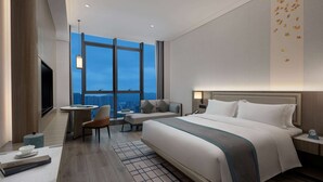 Quarto de hotel moderno com uma cama grande, uma mesa de trabalho, uma cadeira e vista para a cidade através de amplas janelas.