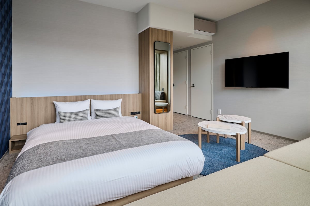 Quarto de hotel moderno com uma cama grande, mesa de cabeceira, televisão e pequena escrivaninha com cadeira.