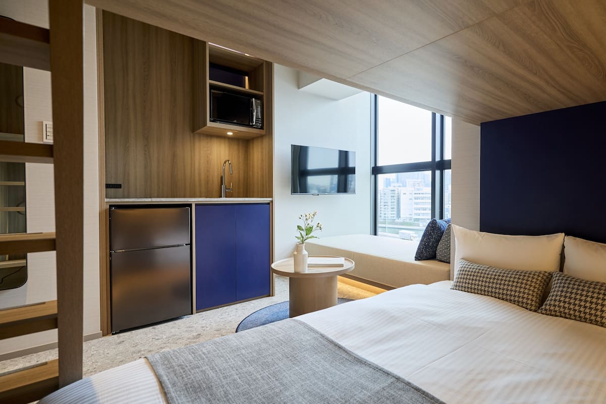Quarto de hotel moderno com cama, cozinha planejada, mesa redonda pequena e uma grande janela com vista para a cidade.