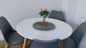 Mesa de jantar redonda com superfície branca e uma rosa rosa solitária num vaso dourado, rodeada por quatro cadeiras cinzas estofadas com pernas de madeira.