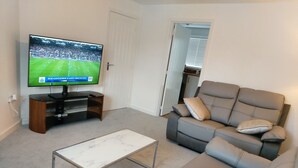 Uma sala de estar com televisão de tela plana, sofá cinza, mesa de centro e uma porta que dá para uma cozinha.