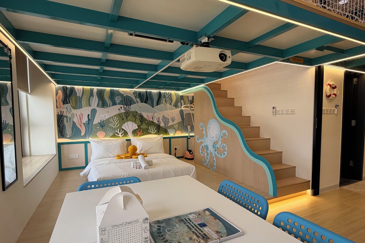 Quarto de hotel moderno com uma cama grande, uma escrivaninha com um guia de boas-vindas, uma escada com detalhes em azul e um projetor no teto.