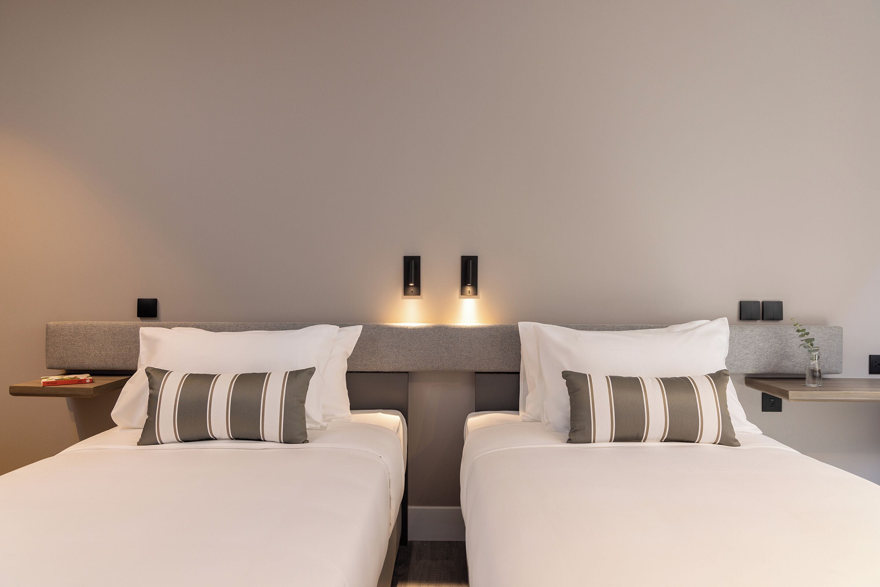 Quarto de hotel com duas camas, uma mesinha de cabeceira e uma luminária fixada na parede.