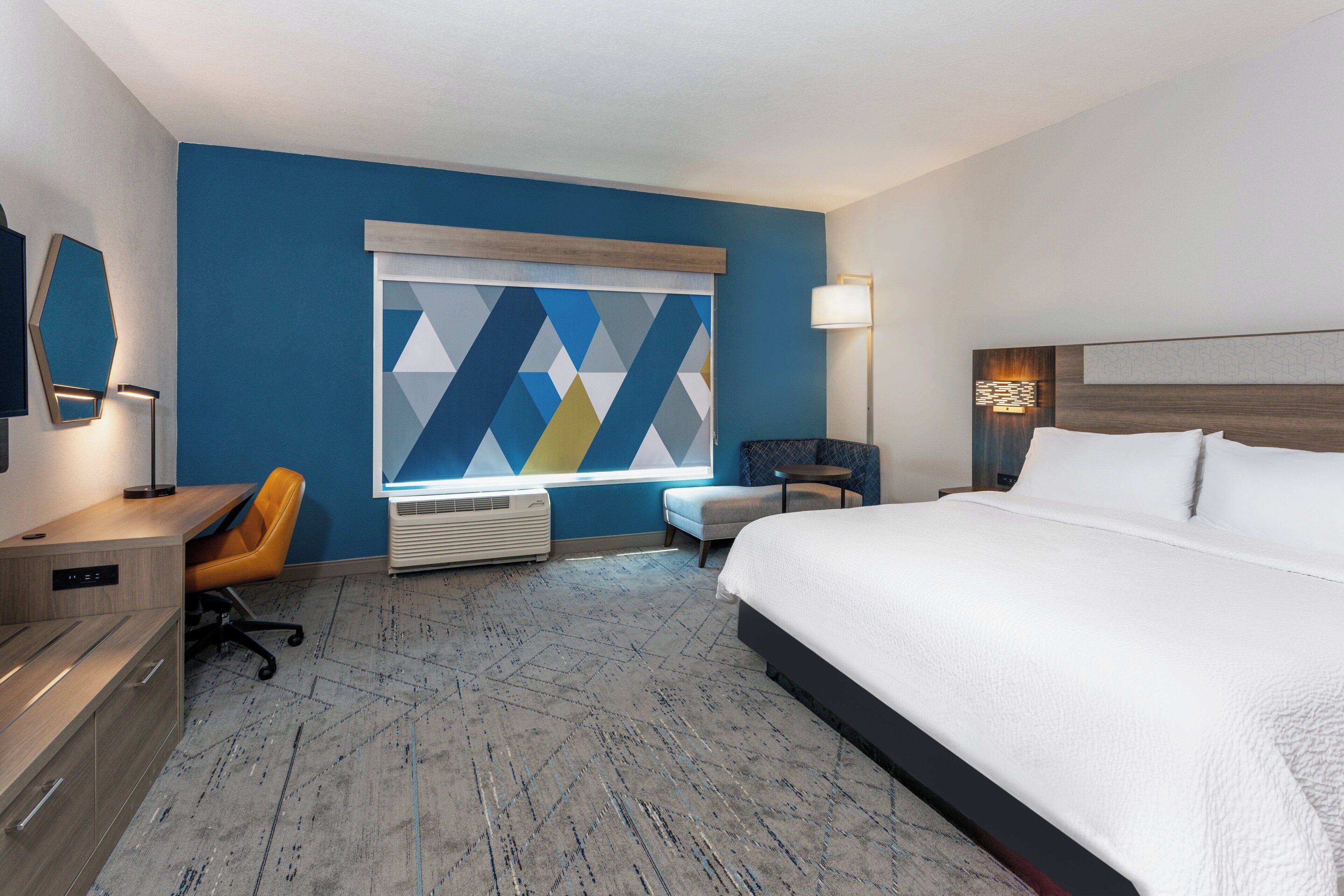 Quarto de hotel com uma cama grande, uma escrivaninha com cadeira, uma janela com vista geométrica e uma luminária fixada na parede.