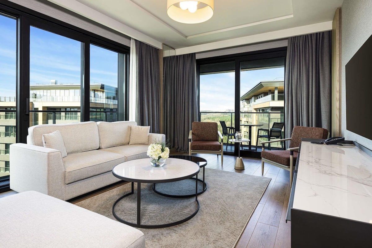 Quarto de hotel moderno com sofá, duas poltronas, mesa de centro e escrivaninha. Grandes janelas proporcionam vista para a cidade.