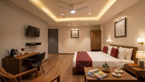 Quarto de hotel com cama, escrivaninha, cadeira, televisão, ventoinha no teto e mesa de cabeceira com abajur.