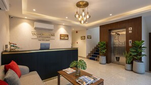 Área de recepção de hotel com balcão, laptop, decoração com bandeira, escada e plantas em vasos.