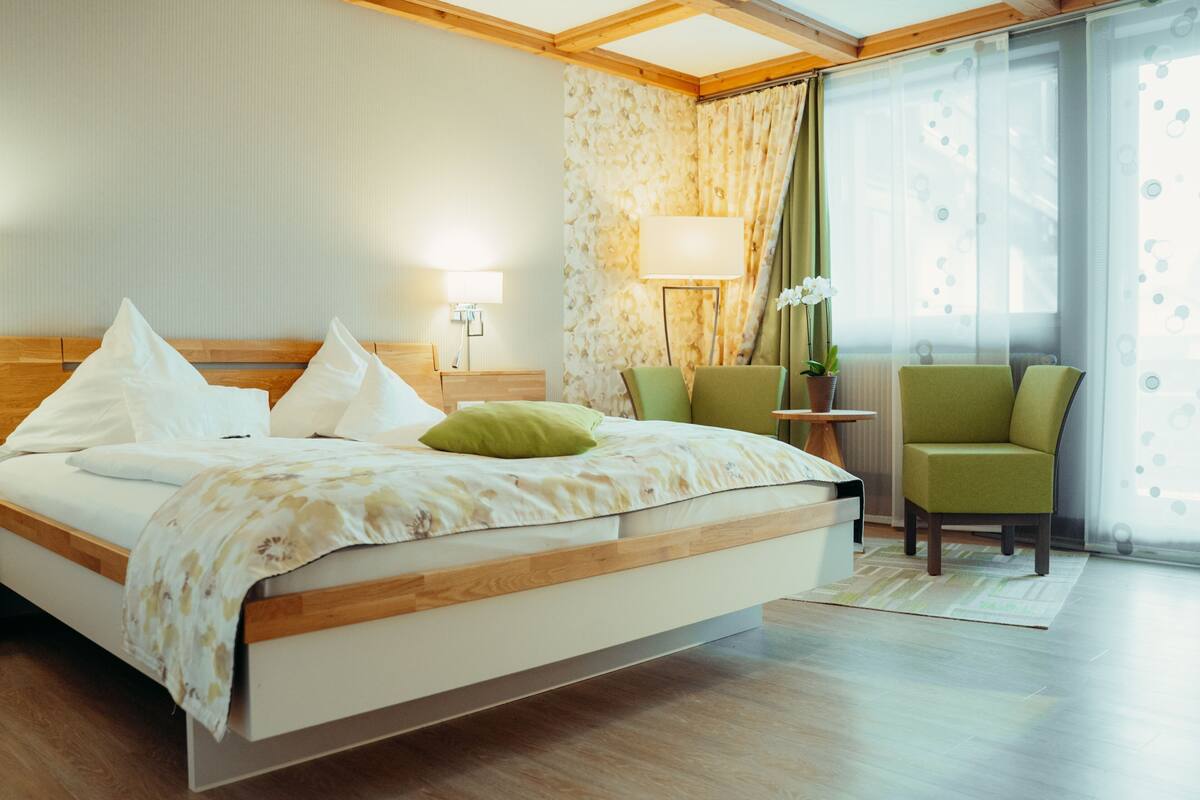 Quarto de hotel moderno com cama de madeira, uma poltrona verde, uma mesinha e uma planta em vaso.