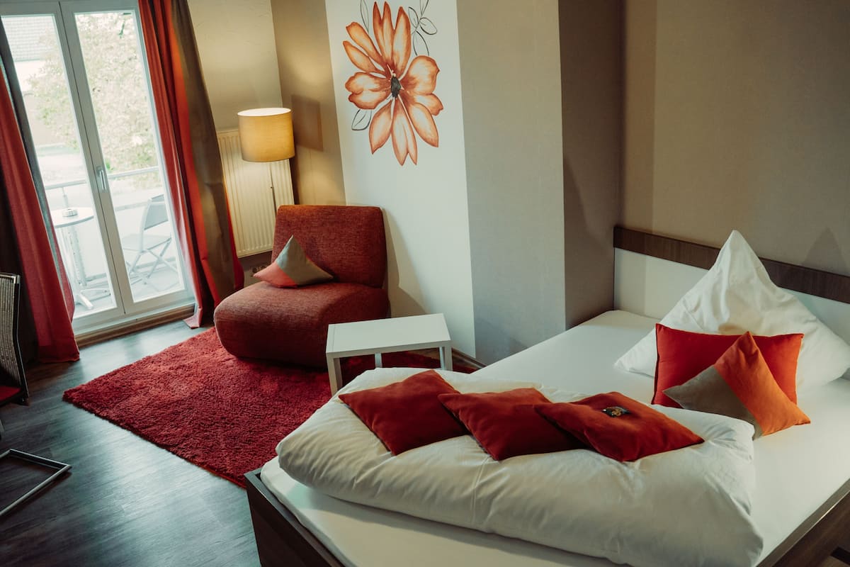 Quarto de hotel com cama, almofadas vermelhas e brancas, uma cadeira vermelha, uma pequena mesa branca e um painel de arte com flores.
