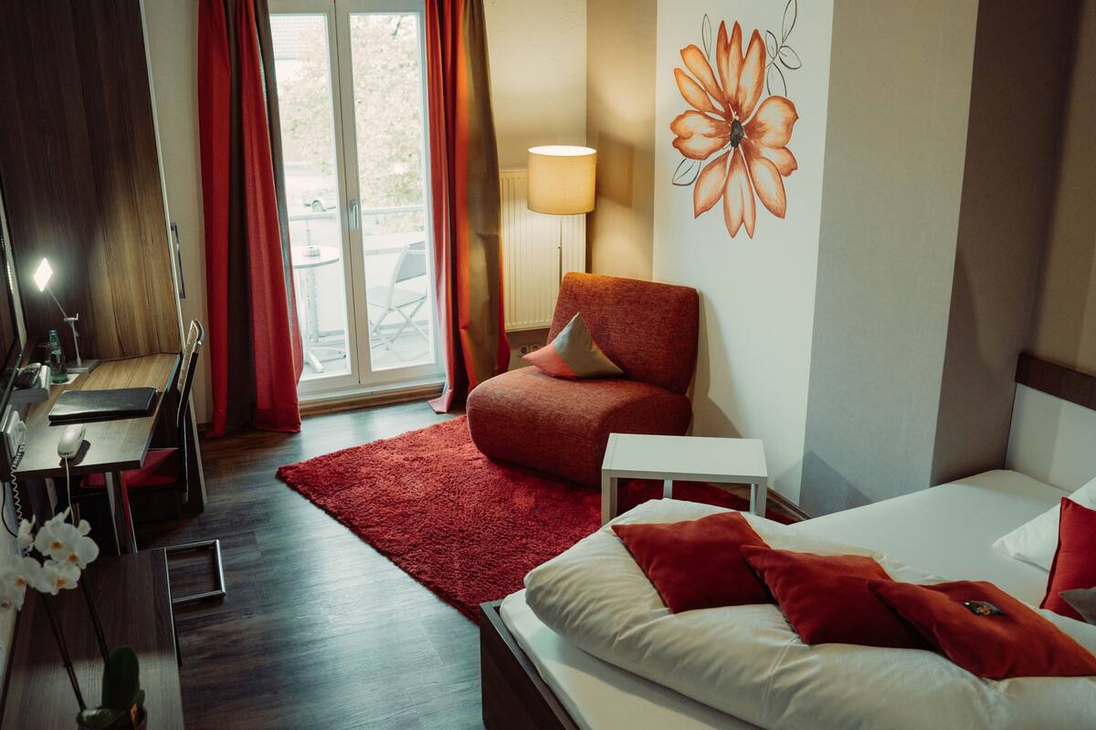 Quarto de hotel com cama, escrivaninha com cadeira, tapete vermelho, poltrona vermelha, mesa de cabeceira branca e um grande mural floral na parede.
