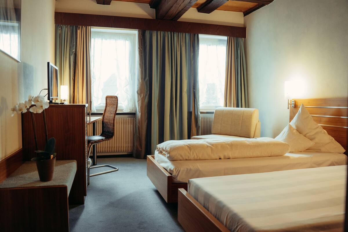 Quarto de hotel com duas camas, uma escrivaninha com cadeira, televisão e janela com cortinas.