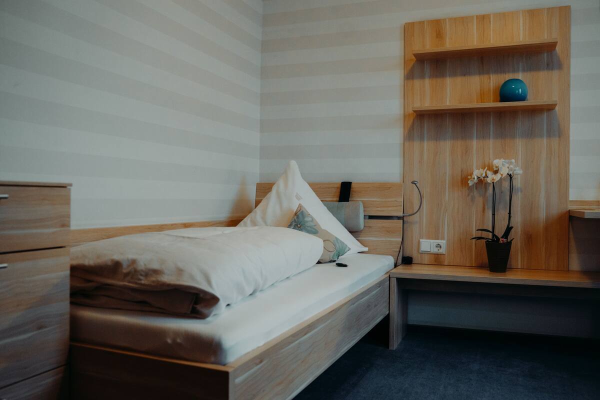 Quarto moderno com cama de madeira, mesa de cabeceira, criado-mudo e prateleira na parede com um vaso e uma planta.