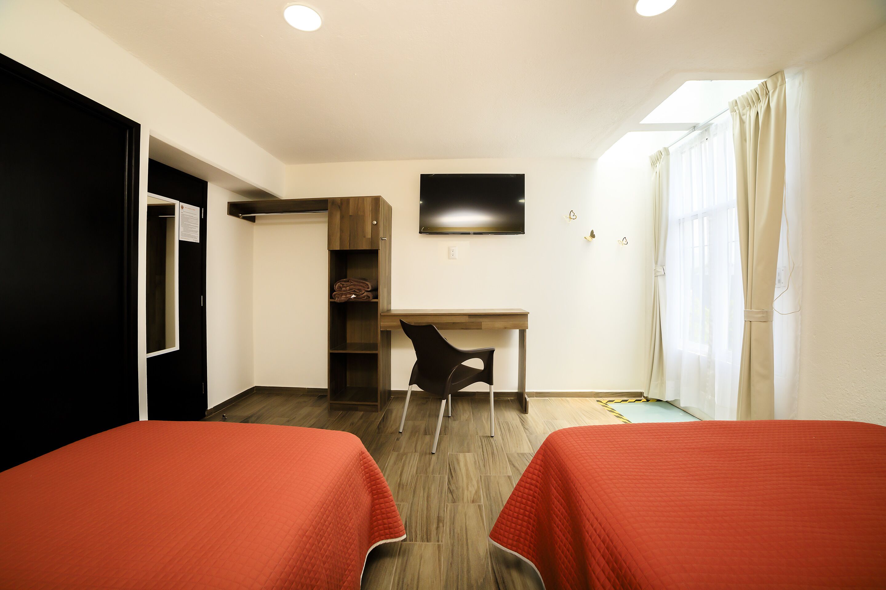 Quarto com duas camas, uma escrivaninha, uma cadeira, uma televisão e janela com cortinas.
