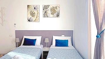 Quarto de hotel com duas camas de solteiro, cada uma com colcha estampada em azul e branco, almofadas brancas e uma almofada decorativa azul. Há duas obras de arte fixadas na parede com padrões de folhas abstratas. O quarto tem um design simples e limpo, com uma paleta de cores neutras.