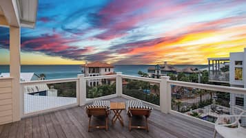 Um deck de madeira com duas cadeiras e uma mesa, com vista para a costa, edifícios e um pôr do sol.