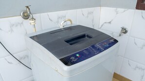 Máquina de lavar compacta, branca e fixada na parede, com painel de controle, localizada num canto com paredes e chão revestidos de azulejos.