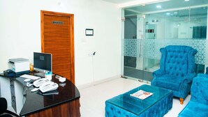 Uma área de recepção com uma porta de madeira com a inscrição 'WASH ROOM', um computador de mesa, um telefone e uma mesa de centro de vidro.