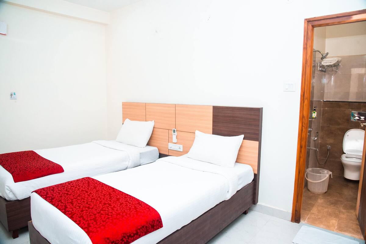 Quarto de hotel com duas camas, cabeceira de madeira, área de chuveiro e vaso sanitário branco.