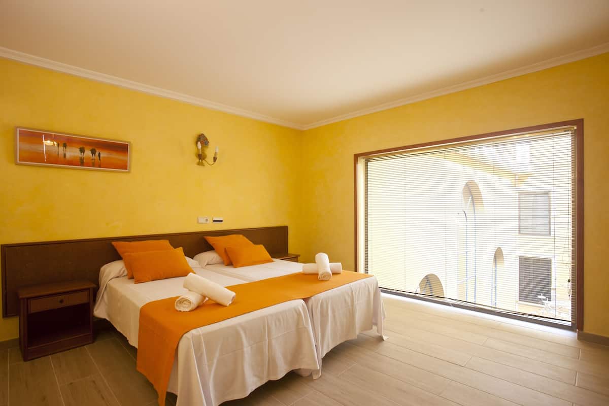 Quarto de hotel com uma cama grande, duas almofadas, uma mesa de cabeceira, um quadro na parede e uma janela com persianas.