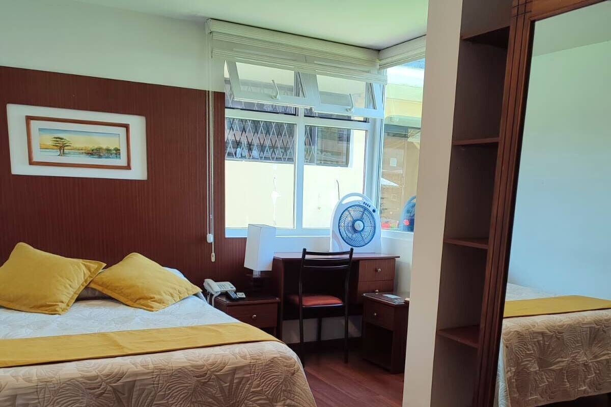 Quarto de hotel com cama, escrivaninha, cadeira, ventoinha e um espelho grande.