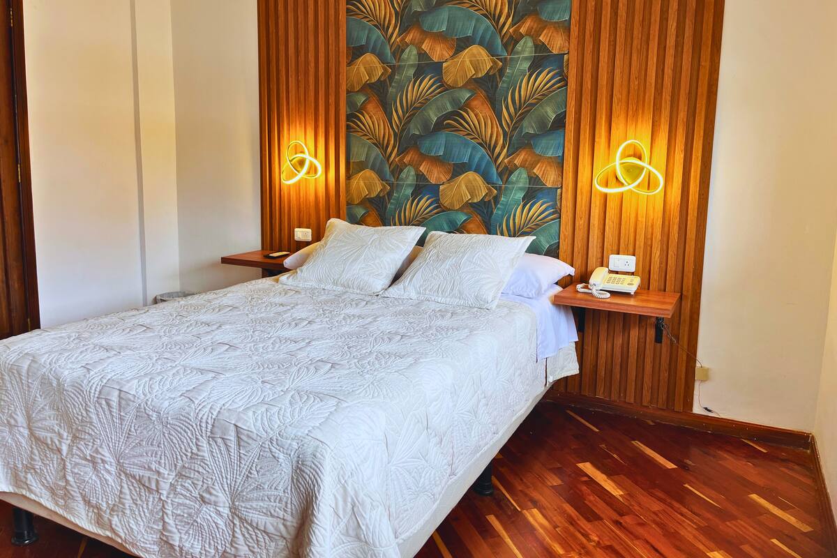 Cama bem arrumada com colcha estampada, cabeceira de madeira e papel de parede com padrão de folhas, além de abajures com luminárias iluminadas.