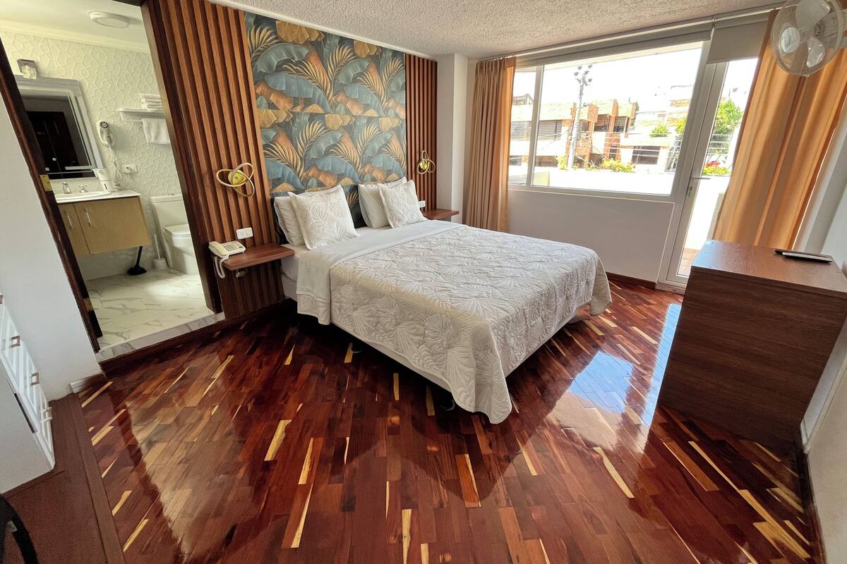 Quarto com cama, soalho de madeira, banheiro com vaso sanitário e janela com cortinas.