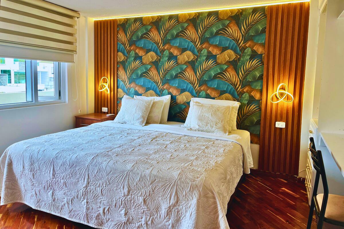 Quarto com cama grande, cabeceira de madeira e parede estampada com folhas tropicais.