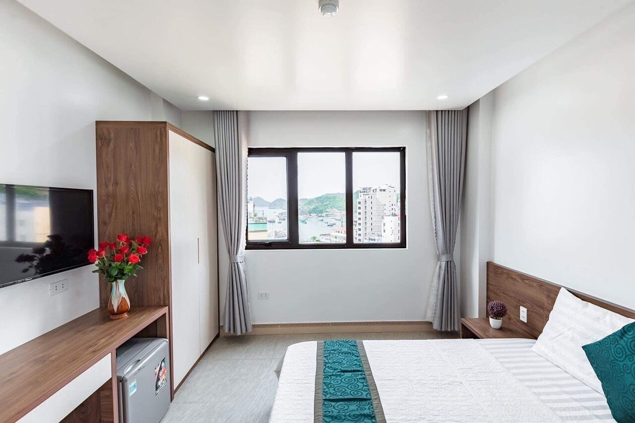 Quarto de hotel com cama, televisão, guarda-roupa de madeira, janela com vista para a cidade e uma pequena mesa com uma planta.