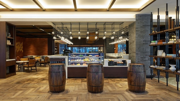 Interior moderno de restaurante com piso de madeira, balcão central para exposição de alimentos e área de estar com mesas e cadeiras.