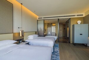 Quarto de hotel com três camas, um espelho grande, um guarda-roupa e um banheiro visível através de uma porta de vidro.