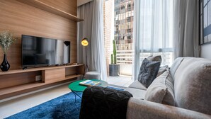 Uma sala de estar moderna com televisão de tela plana, prateleiras de madeira, mesa de centro verde, sofá e uma janela grande com vista para um edifício.