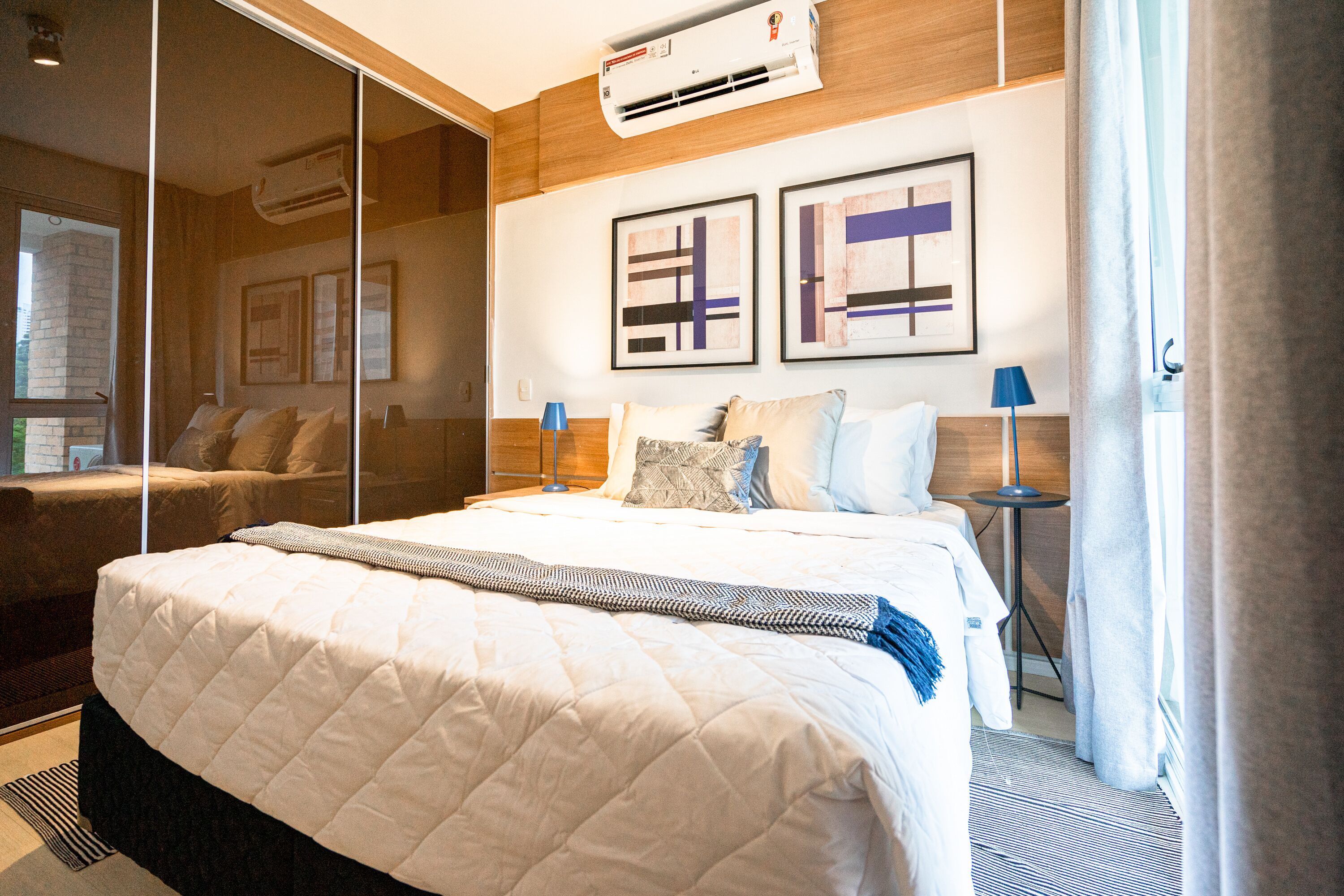 Quarto de hotel moderno com uma cama grande, mesa de cabeceira com abajur, ar condicionado fixado na parede e arte abstrata na parede.